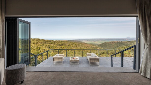 Uma varanda com vista para uma paisagem montanhosa, uma mesa de jantar com arranjo de flores e um espaço de estar com almofadas.