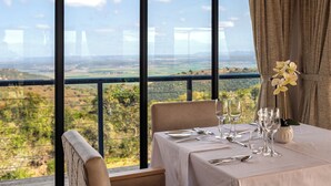 Mesa de jantar posta com porcelana fina, talheres e taças de vinho, com vista para uma paisagem cênica com vegetação e construções ao longe.