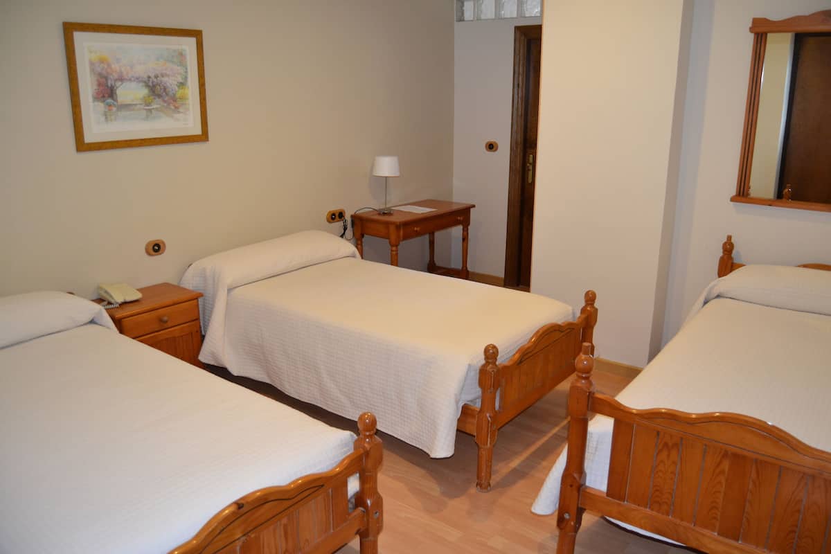 Quarto de hotel com três camas de madeira, uma mesinha de cabeceira com abajur, um quadro na parede e uma porta que dá para outro cômodo.
