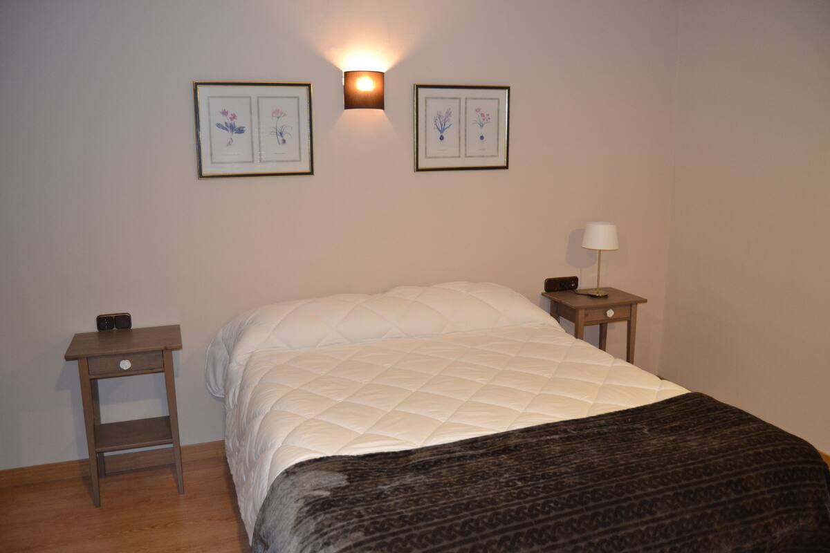 Um quarto com uma cama, uma mesinha de cabeceira e duas gravuras de plantas emolduradas na parede.