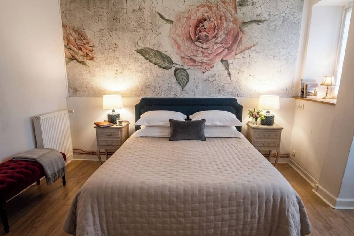 Quarto com uma cama grande, duas mesas de cabeceira com luminárias, um radiador, um banco e um mural floral na parede.