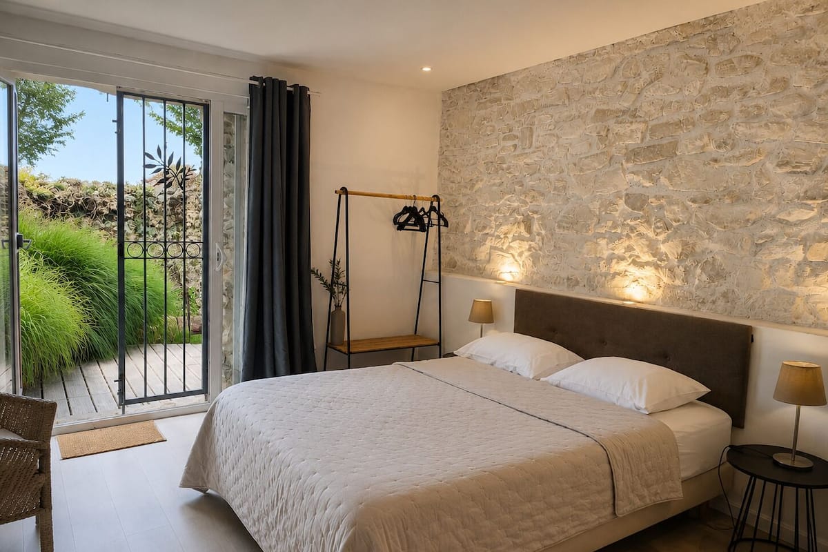 Quarto com cama, duas mesas de cabeceira, guarda-roupa, varanda com vista para o verde e parede de pedra.