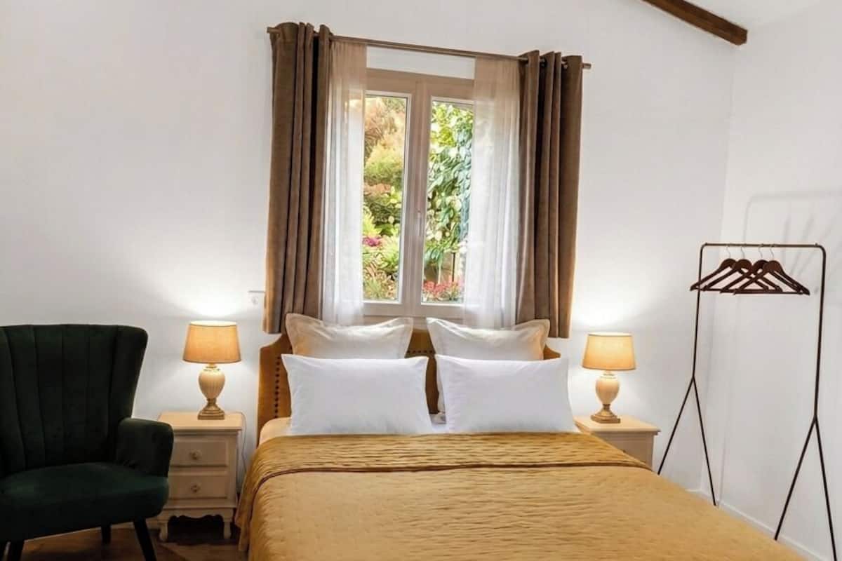 Quarto com cama, duas mesas de cabeceira, uma cadeira, luminária pendente e janela com cortinas.
