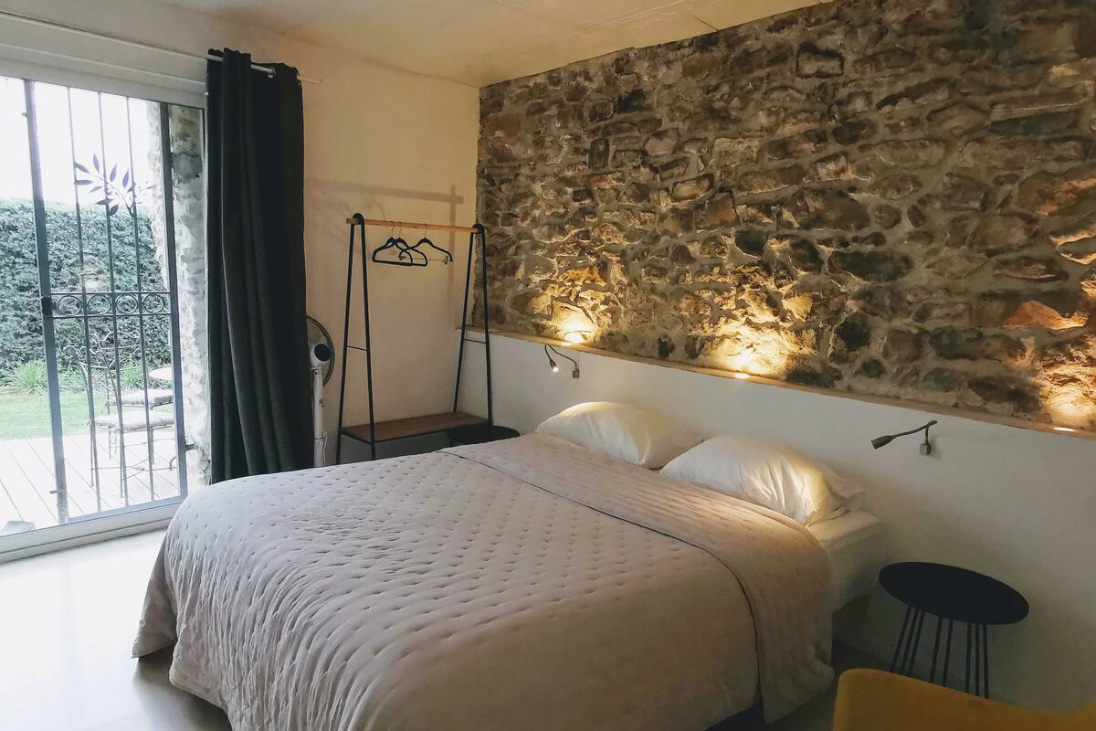 Quarto com parede de pedra, cama com almofadas, cadeira suspensa e uma pequena mesa.