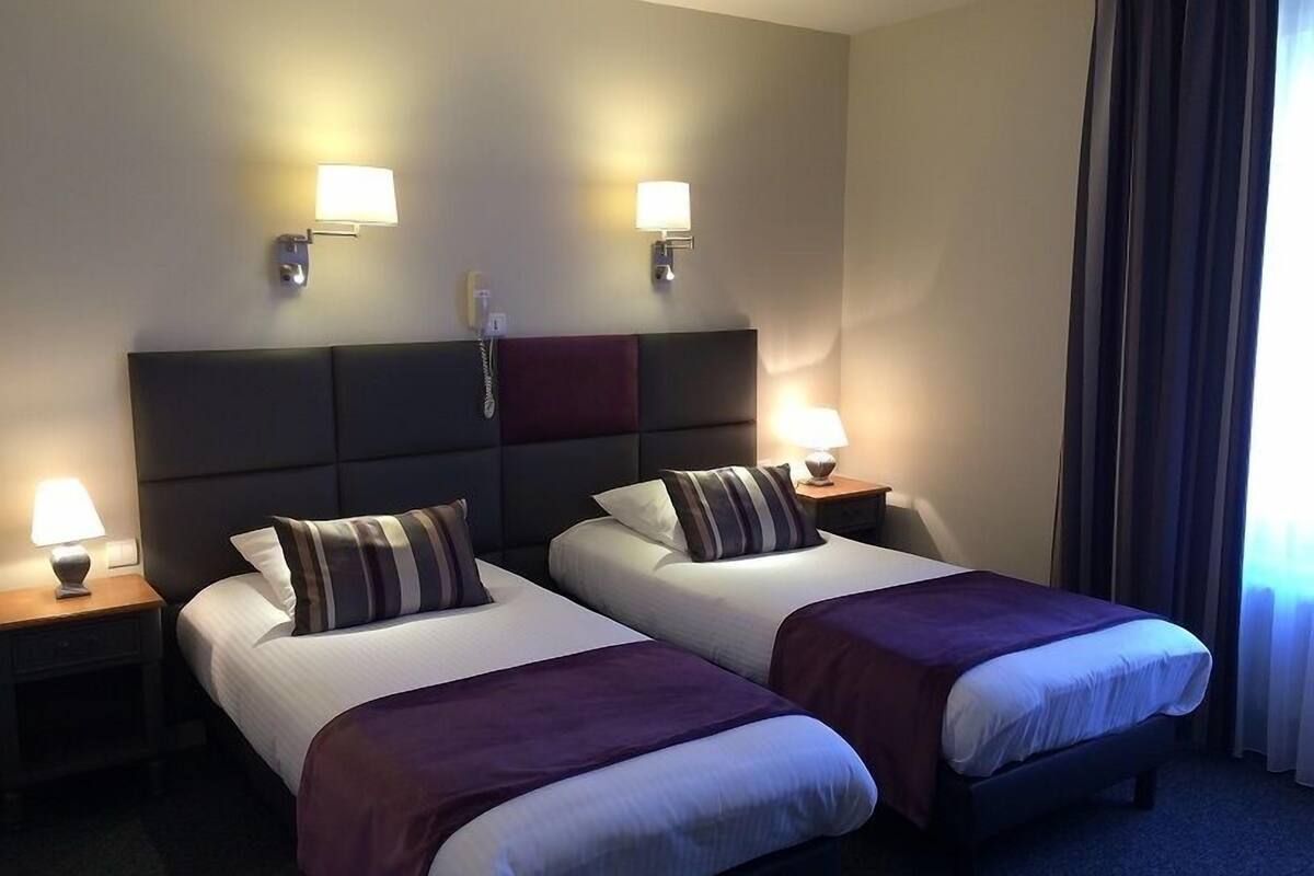 Quarto de hotel com duas camas, cada uma com travesseiro e cobertor. Há mesas de cabeceira com abajures e um telefone na parede.