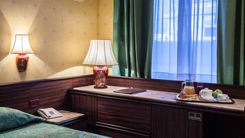 Quarto de hotel com cama, mesa de cabeceira, luminárias, minibar e vista para o exterior através de uma janela com cortinas.