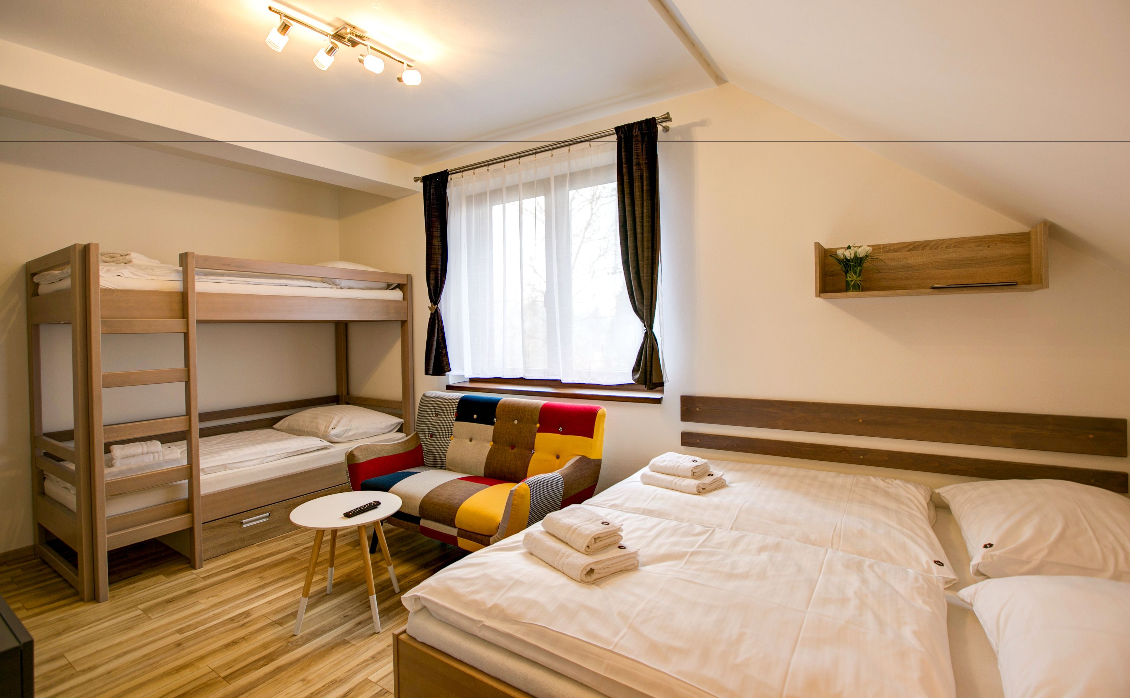 Quarto com beliches, uma cama de solteiro, uma mesinha e uma janela com cortinas.
