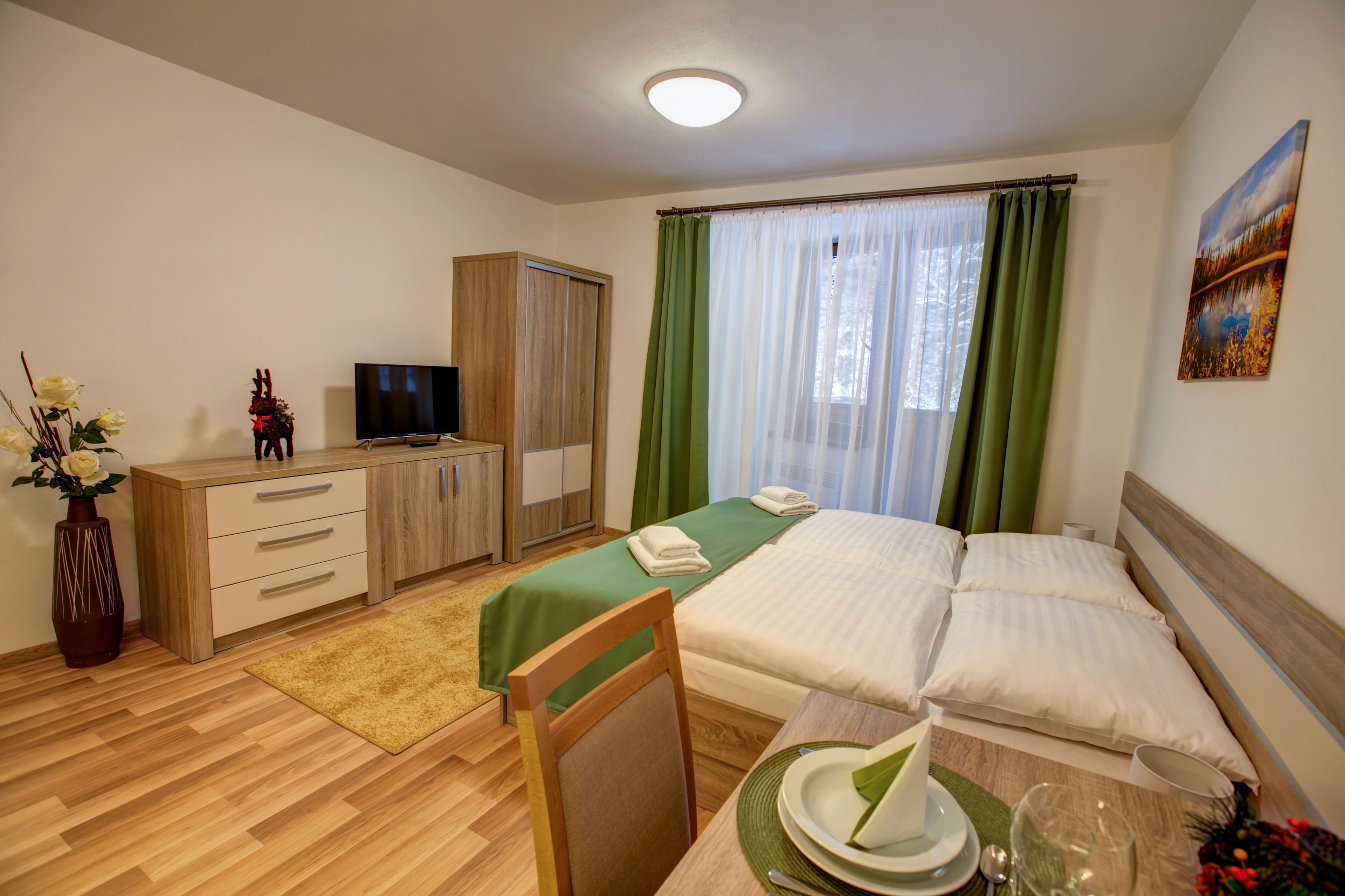 Quarto de hotel com cama, escrivaninha, televisão, guarda-roupa e janela com cortinas.