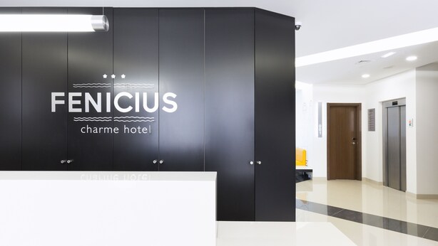 Uma área de recepção de hotel moderna, com um balcão elegante preto e uma placa com a inscrição "FENICIUS charme hotel".
