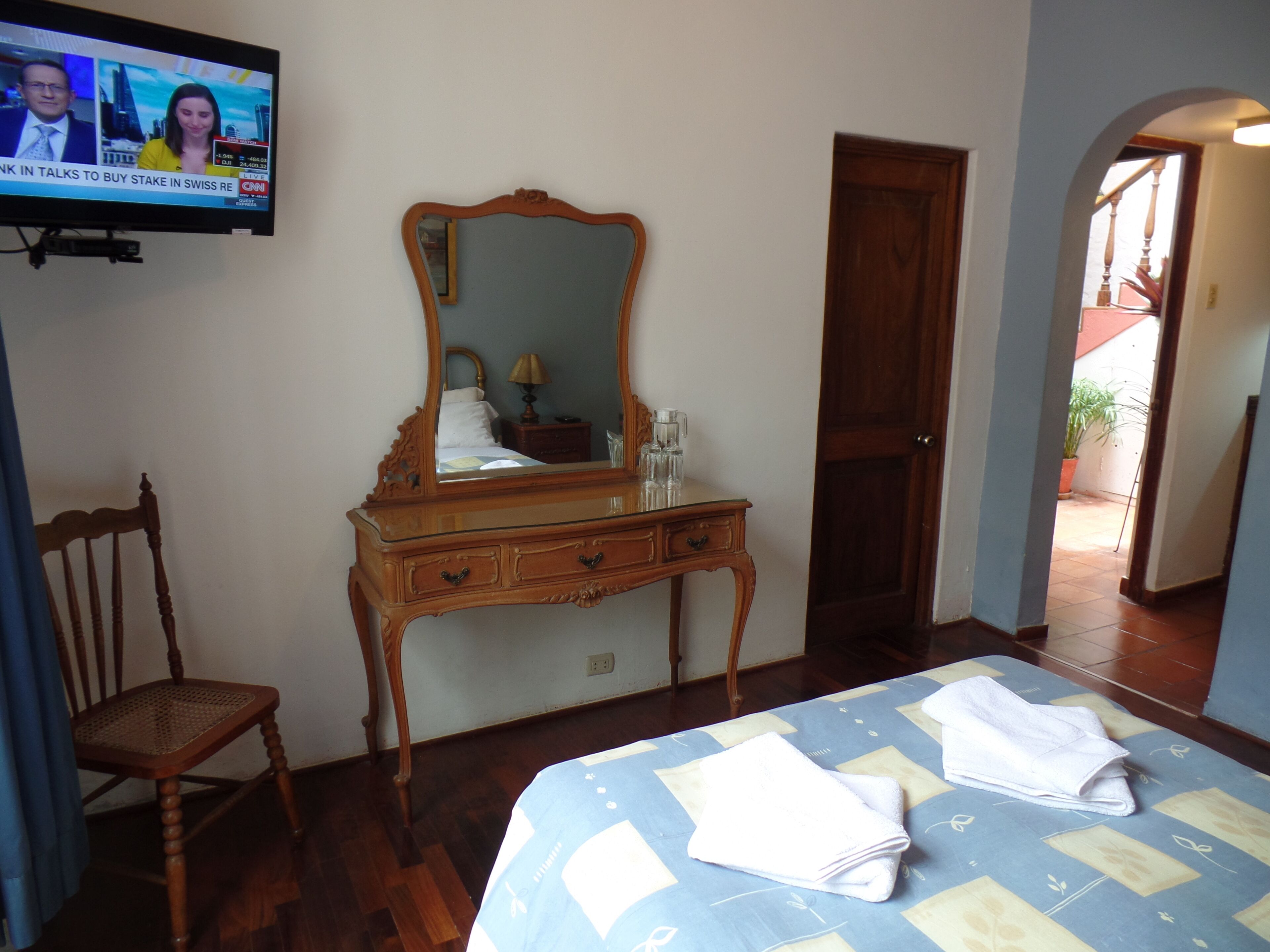 Quarto com cômoda de madeira, uma cadeira, cama com colcha estampada em azul e branco, e uma TV exibindo uma transmissão de notícias.