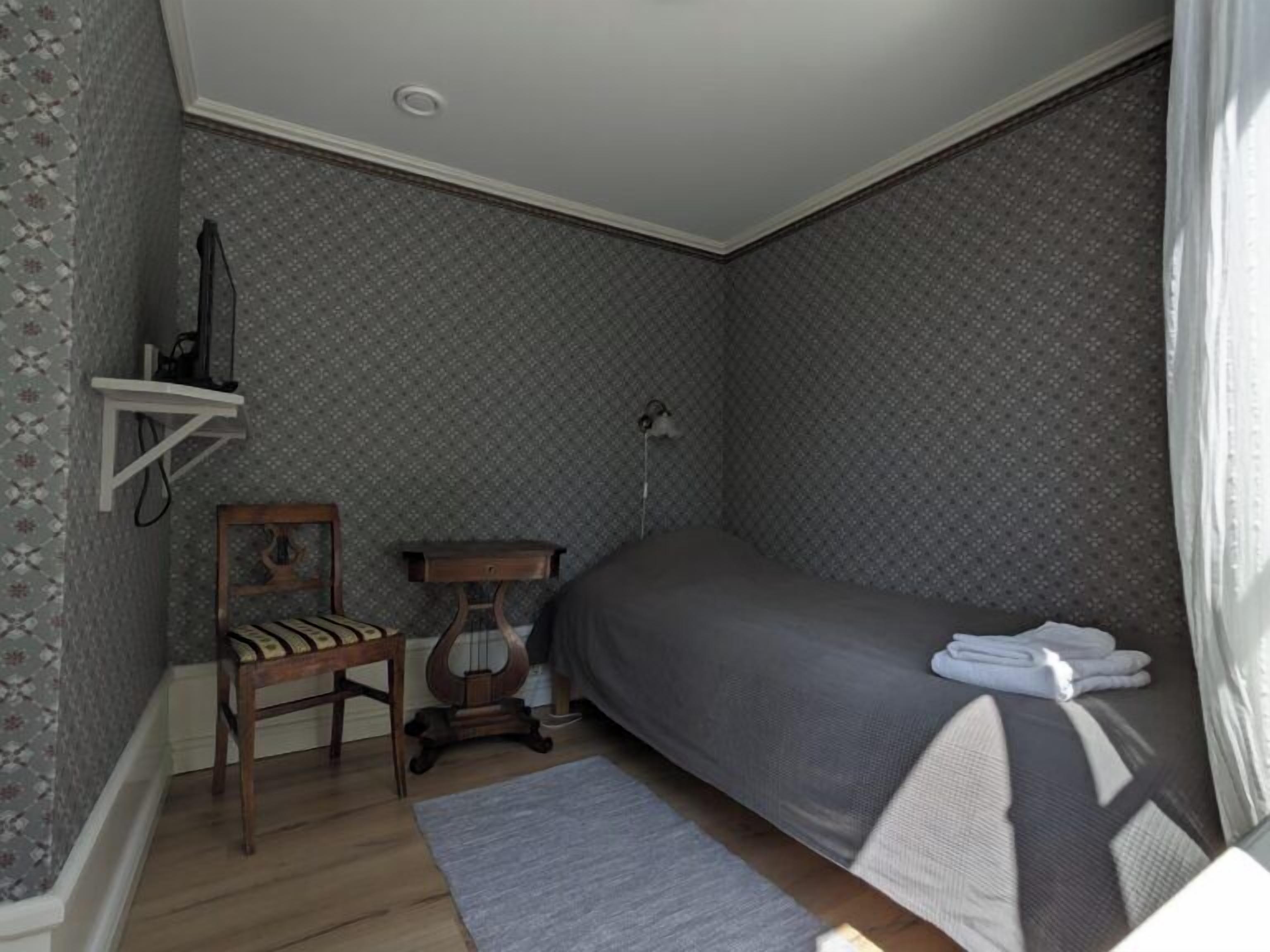 Um quarto pequeno e bem organizado, com uma cama, uma cadeira, uma mesinha e uma prateleira.