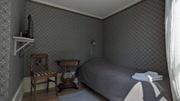 Um quarto pequeno e bem organizado, com uma cama, uma cadeira, uma mesinha e uma prateleira.