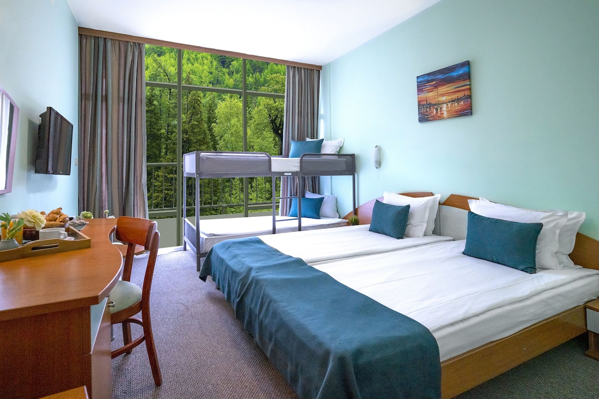 Quarto de hotel com uma cama grande, uma escrivaninha com cadeira, uma beliche e um quadro na parede.