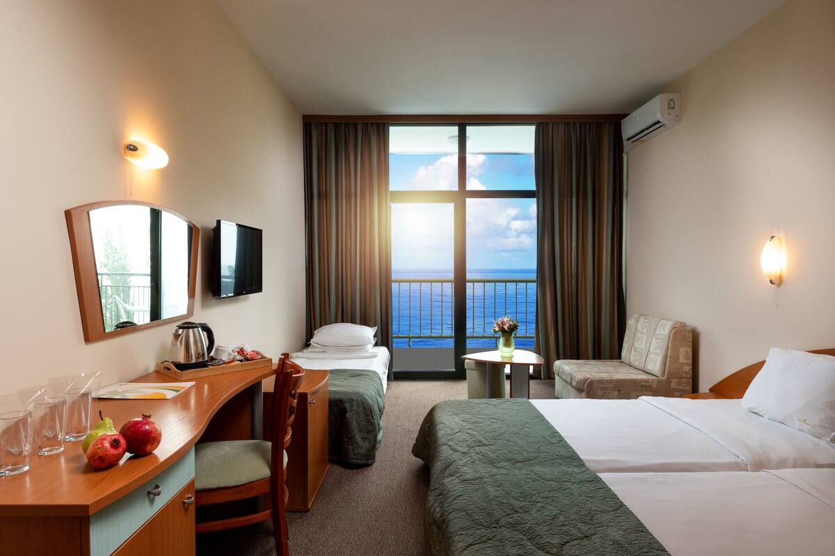 Quarto de hotel com duas camas, uma escrivaninha com cadeira, uma mesinha, televisão e uma grande janela com vista para o mar.