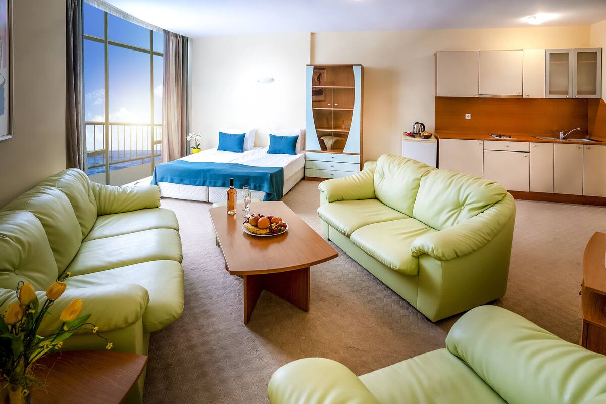 Quarto de hotel com cama, dois sofás verdes, mesa de centro de madeira, área de cozinha e uma janela grande com vista.