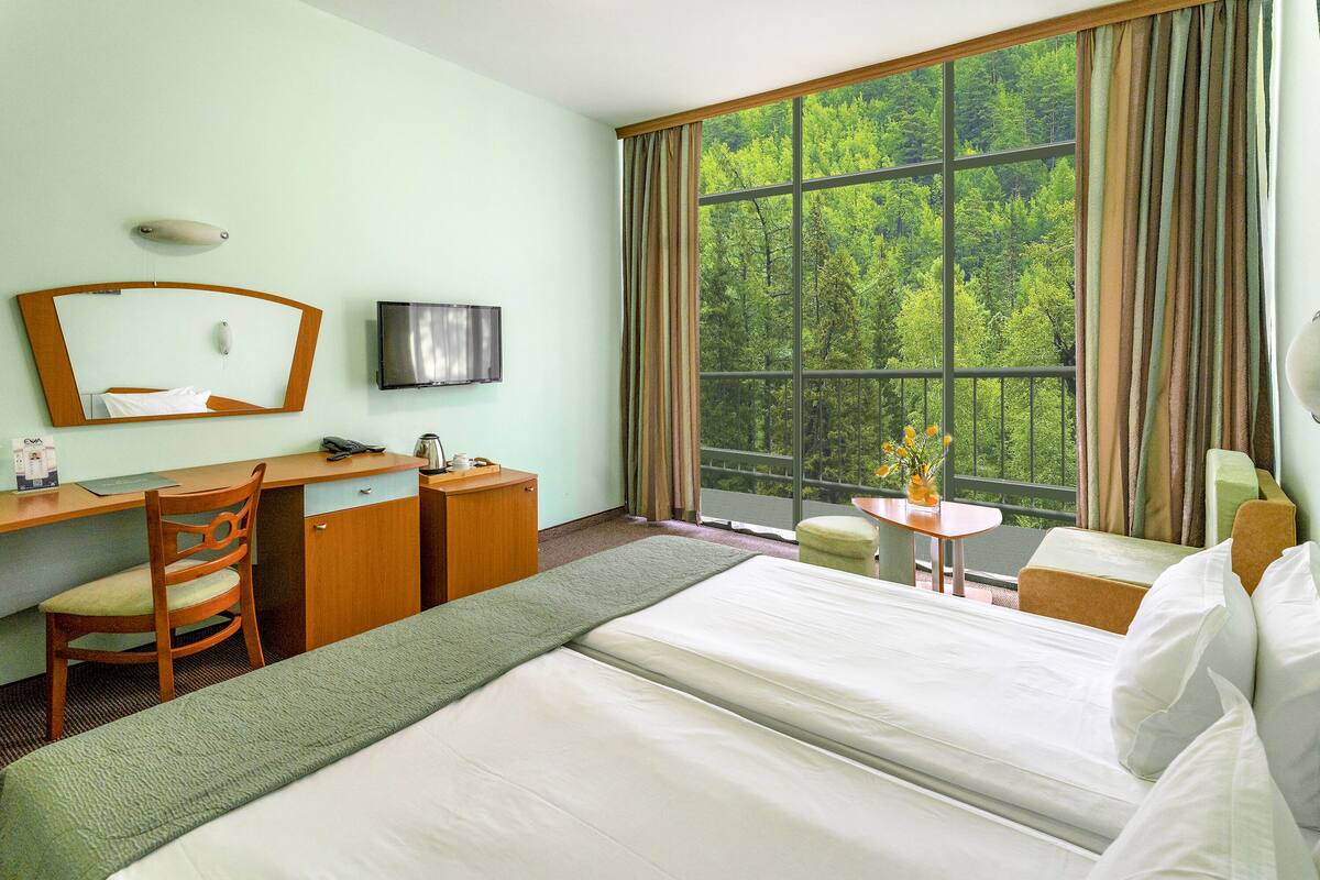 Quarto de hotel com cama, escrivaninha, cadeira, televisão e uma janela grande com vista para árvores.