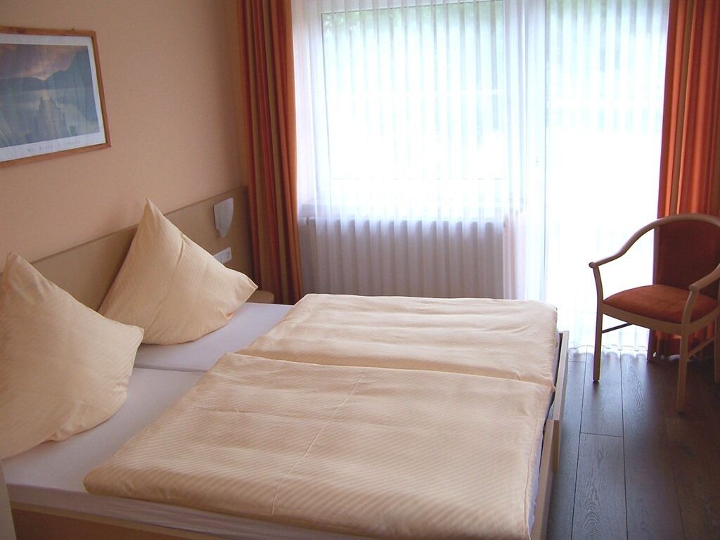 Quarto de hotel com uma cama, duas almofadas, uma cadeira, uma janela com cortinas e um quadro na parede.