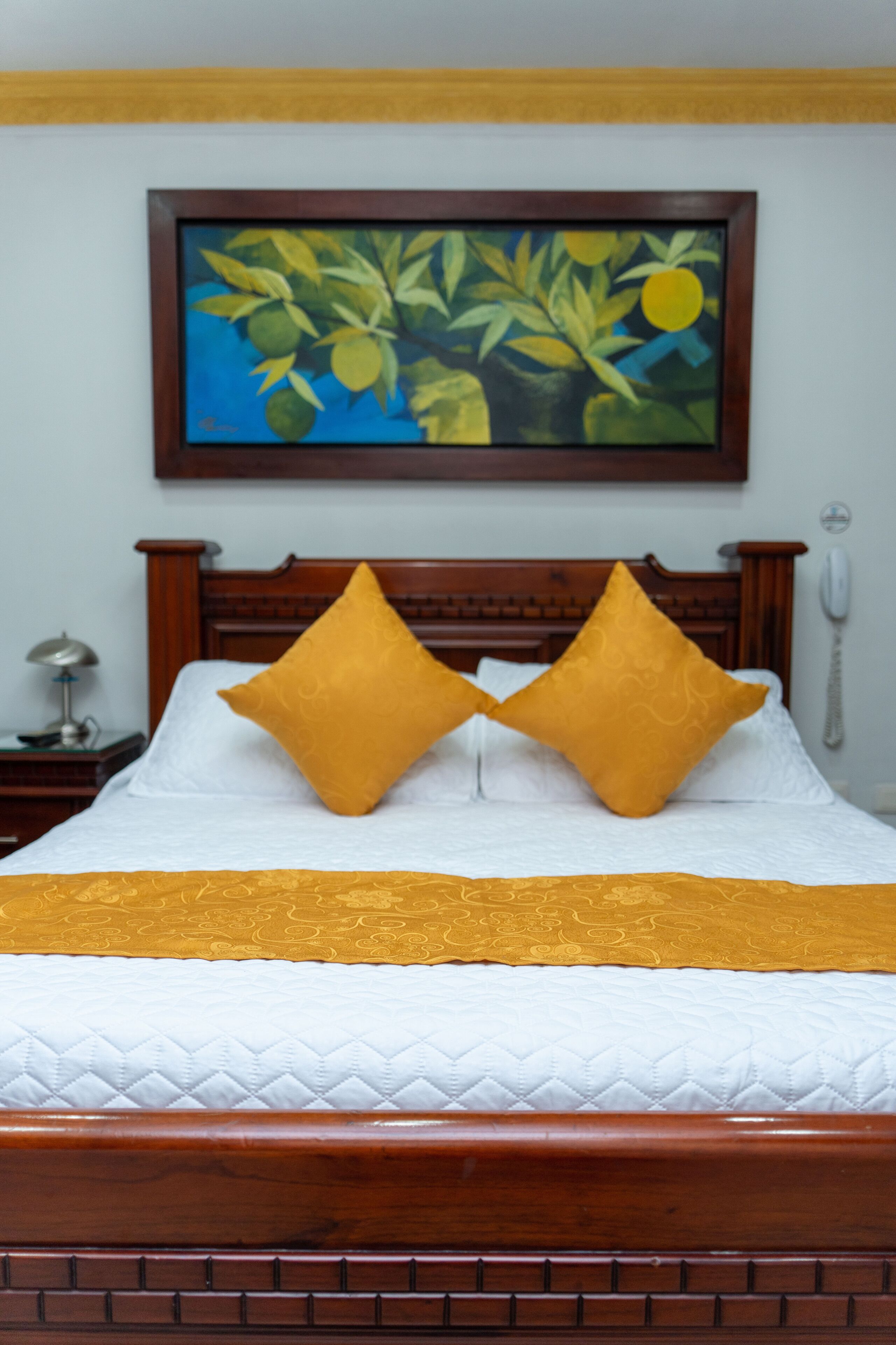Cama bem arrumada com cabeceira de madeira, duas almofadas douradas e colcha branca. Um quadro com folhas verdes e círculos amarelos está pendurado acima da cama.