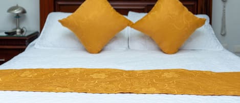 Cama bem arrumada com cabeceira de madeira, duas almofadas douradas e colcha branca. Um quadro com folhas verdes e círculos amarelos está pendurado acima da cama.
