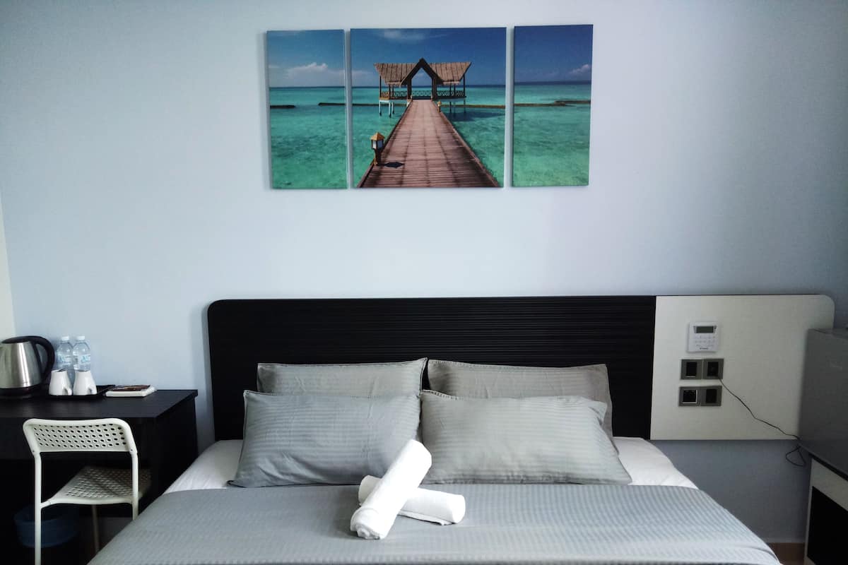 Quarto de hotel com cama, almofadas, escrivaninha, cadeira, mesa de cabeceira, abajur e tomada na parede.