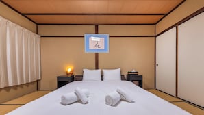Cama bem arrumada com lençóis brancos e toalhas enroladas, mesa de cabeceira com abajur e quadro na parede.