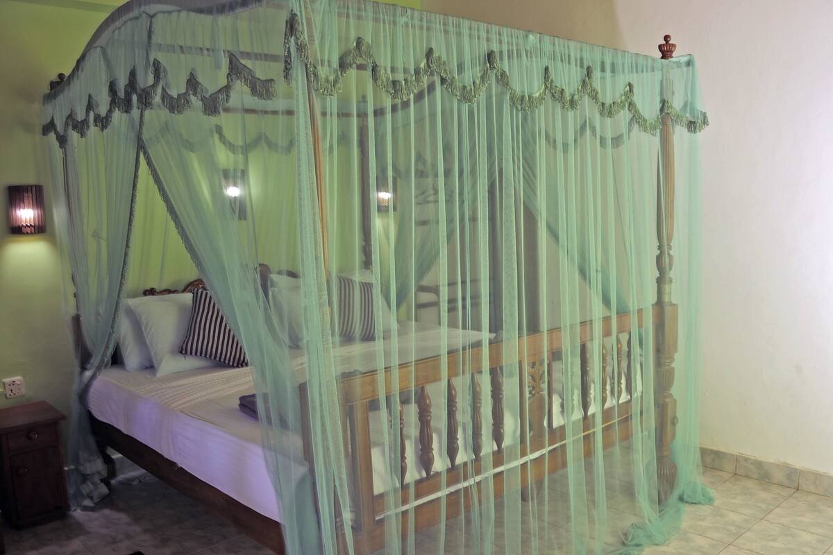 Uma cama com dossel, uma mesinha de cabeceira e um candeeiro fixo na parede.