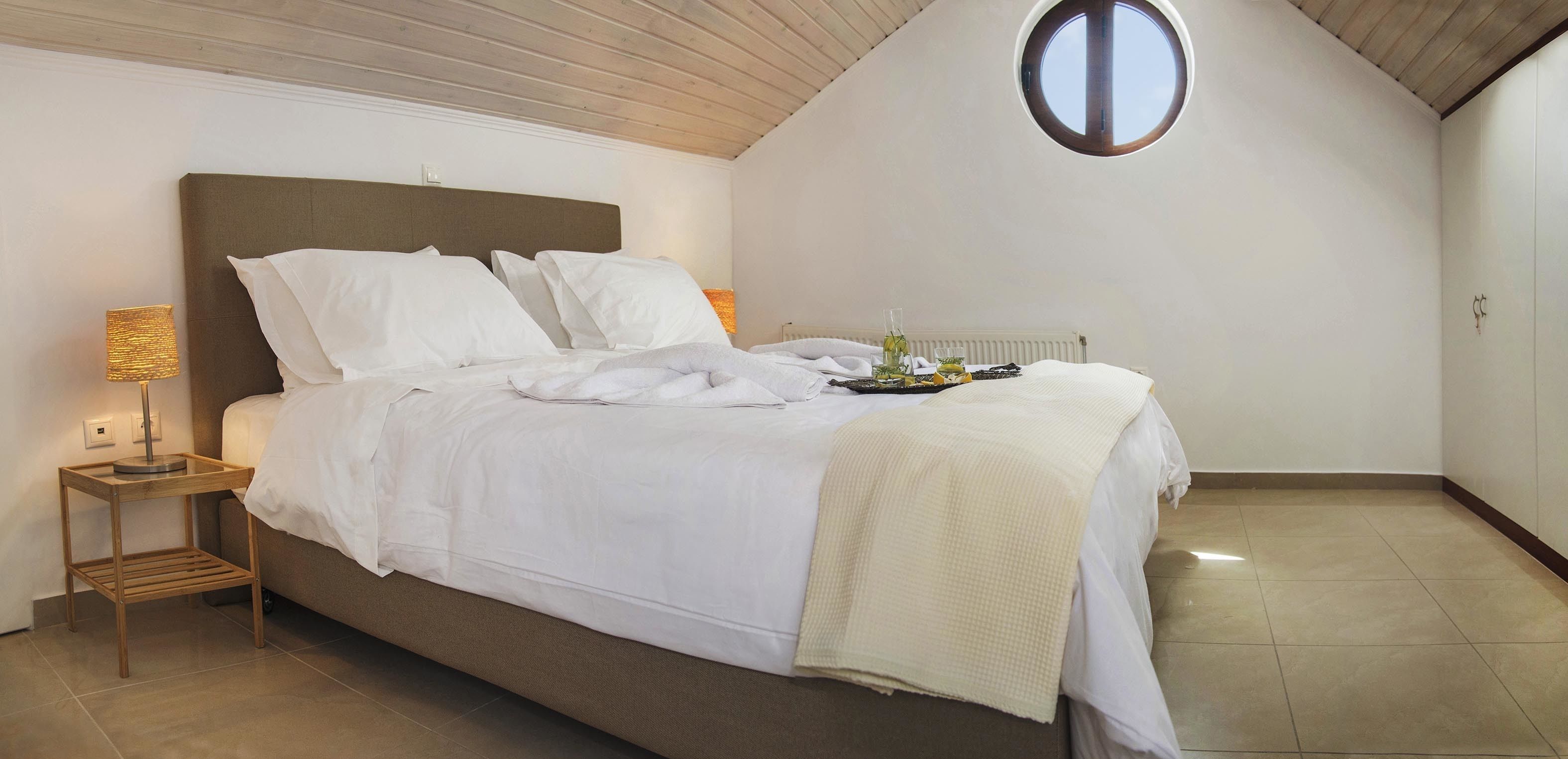 Quarto com cama, mesa de cabeceira, luminária e janela redonda.