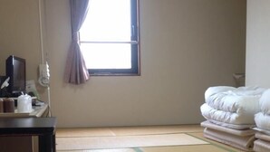 Quarto com tatames, cama com roupa de cama branca, escrivaninha com computador e janela com cortinas.