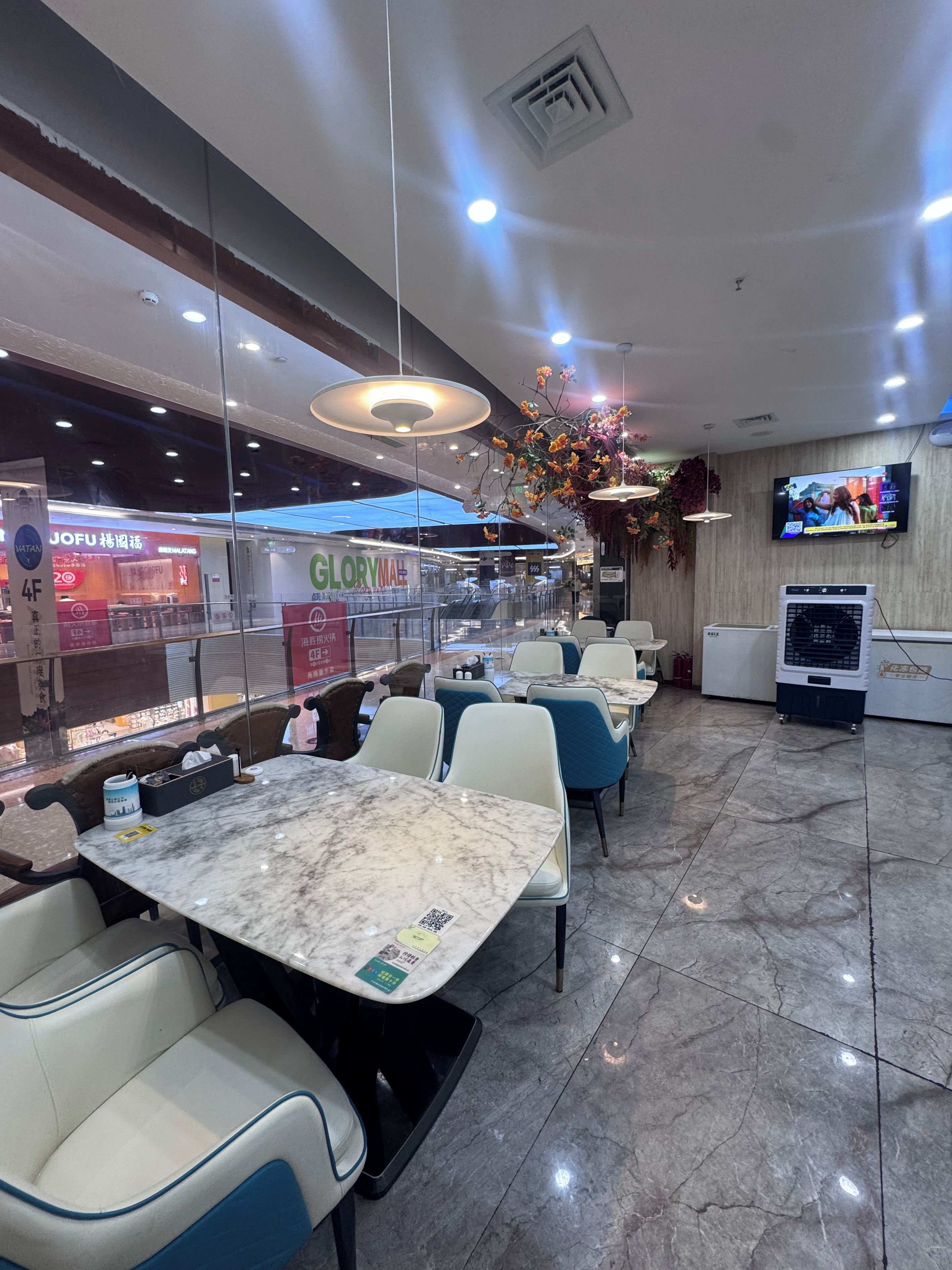 Um restaurante com mesas e cadeiras de mármore, uma tela de televisão e teto com iluminação embutida.