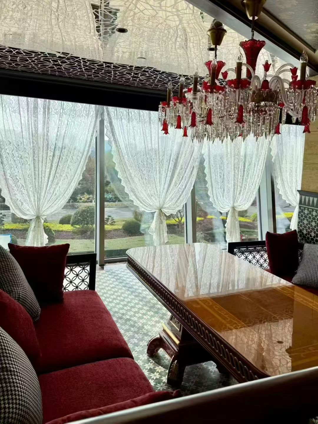 Quarto com grande mesa de jantar, sofás estofados vermelhos e vista para um jardim através de amplas janelas com cortinas brancas.