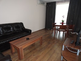 Uma sala de estar com um sofá de couro preto, uma mesa de centro de madeira, uma mesa de jantar com cadeiras e um rack para TV com controle remoto.