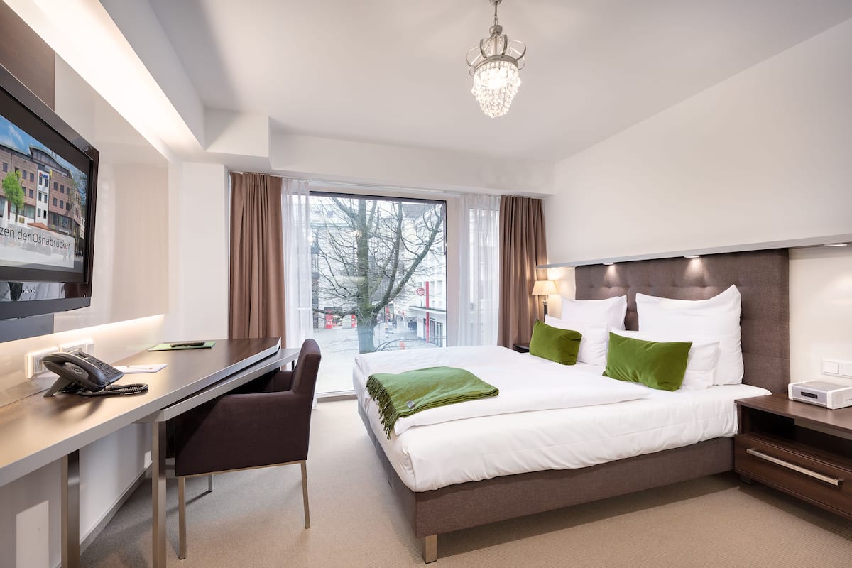 Quarto de hotel com cama, escrivaninha, cadeira e televisão exibindo uma vista da cidade.