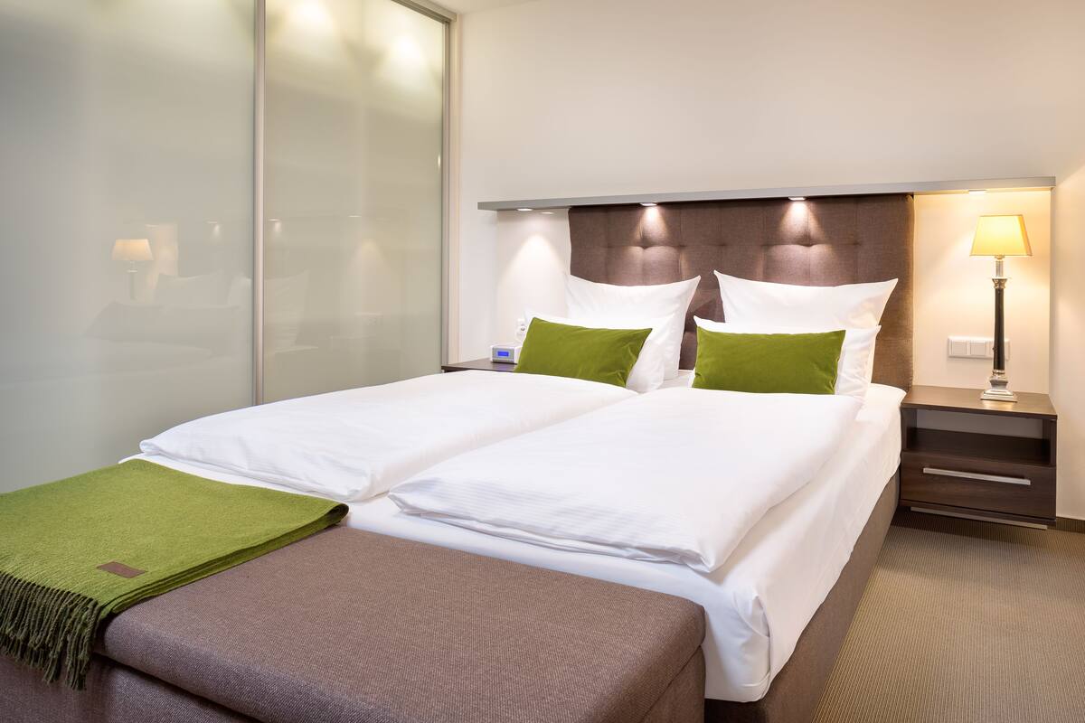 Cama de casal com lençóis brancos e almofadas verdes, mesa de cabeceira com luminária e um banco marrom.