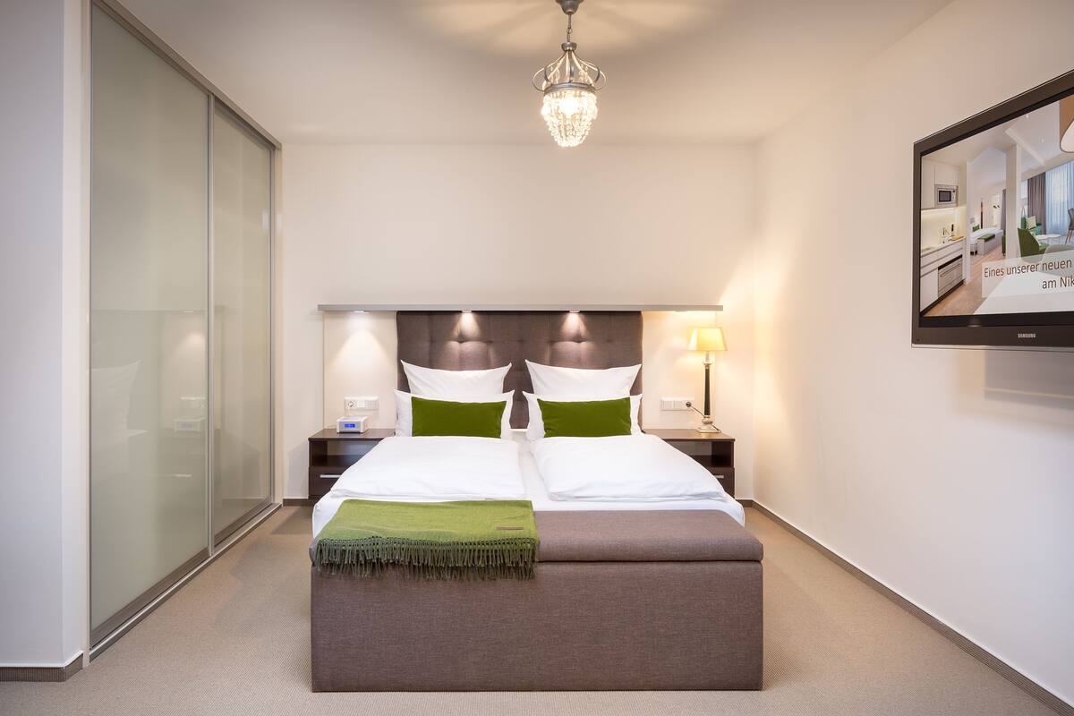 Quarto de hotel moderno com cama grande, televisão de tela plana, mesa de cabeceira e armário com porta de vidro deslizante.