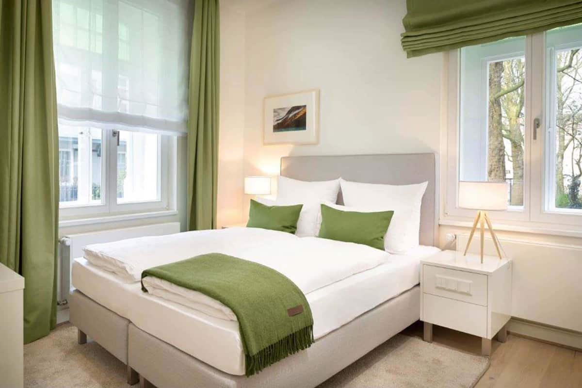 Uma cama bem arrumada com um manta verde, uma mesa de cabeceira com abajur e uma janela com cortinas.