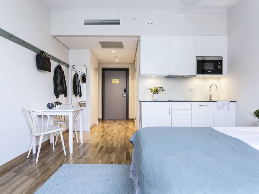 Quarto de hotel moderno com uma cama, uma pequena escrivaninha, uma cadeira, uma cozinha compacta e um banheiro.