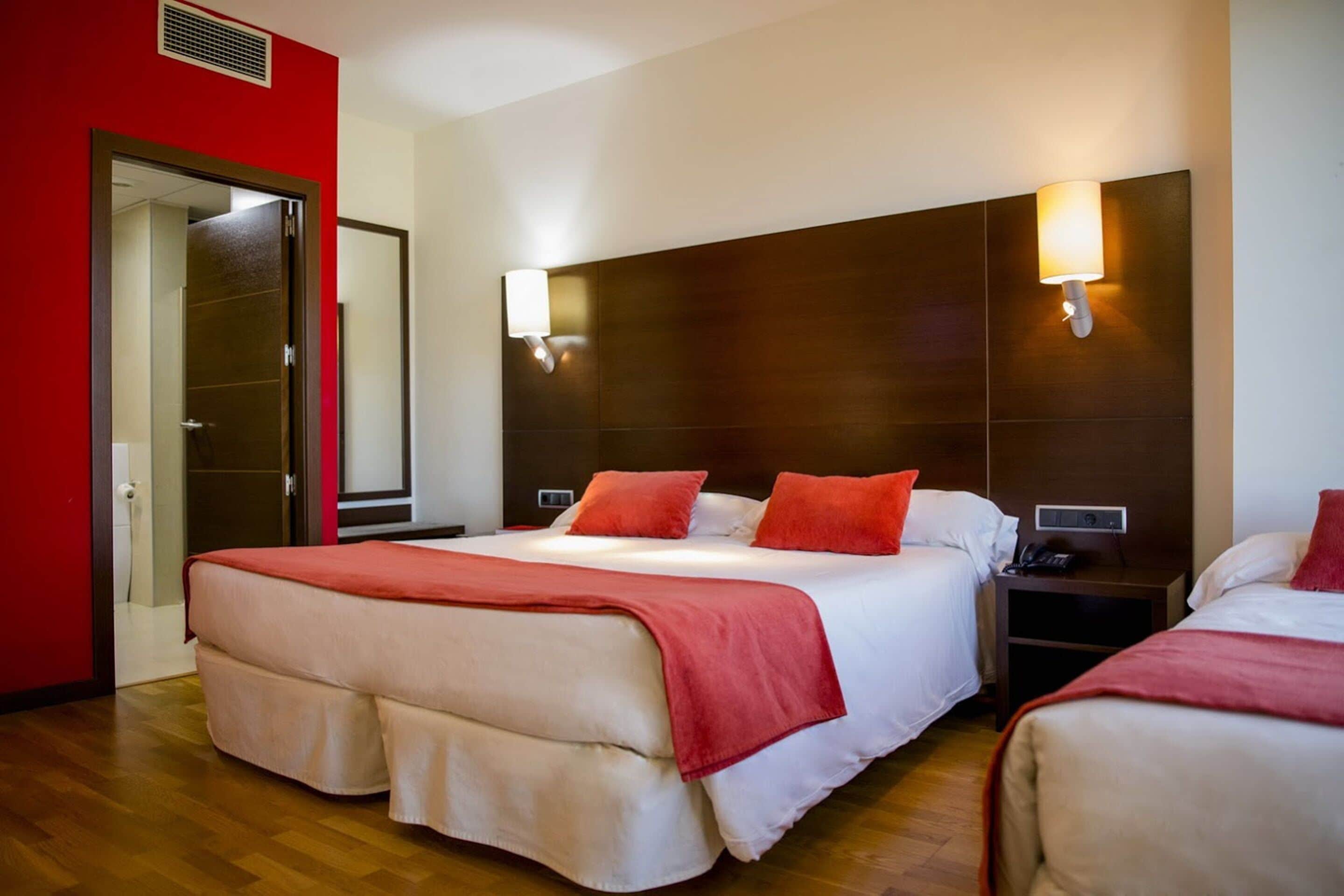 Quarto de hotel com duas camas, detalhes vermelhos, uma cabeceira de madeira e luminárias de cabeceira.