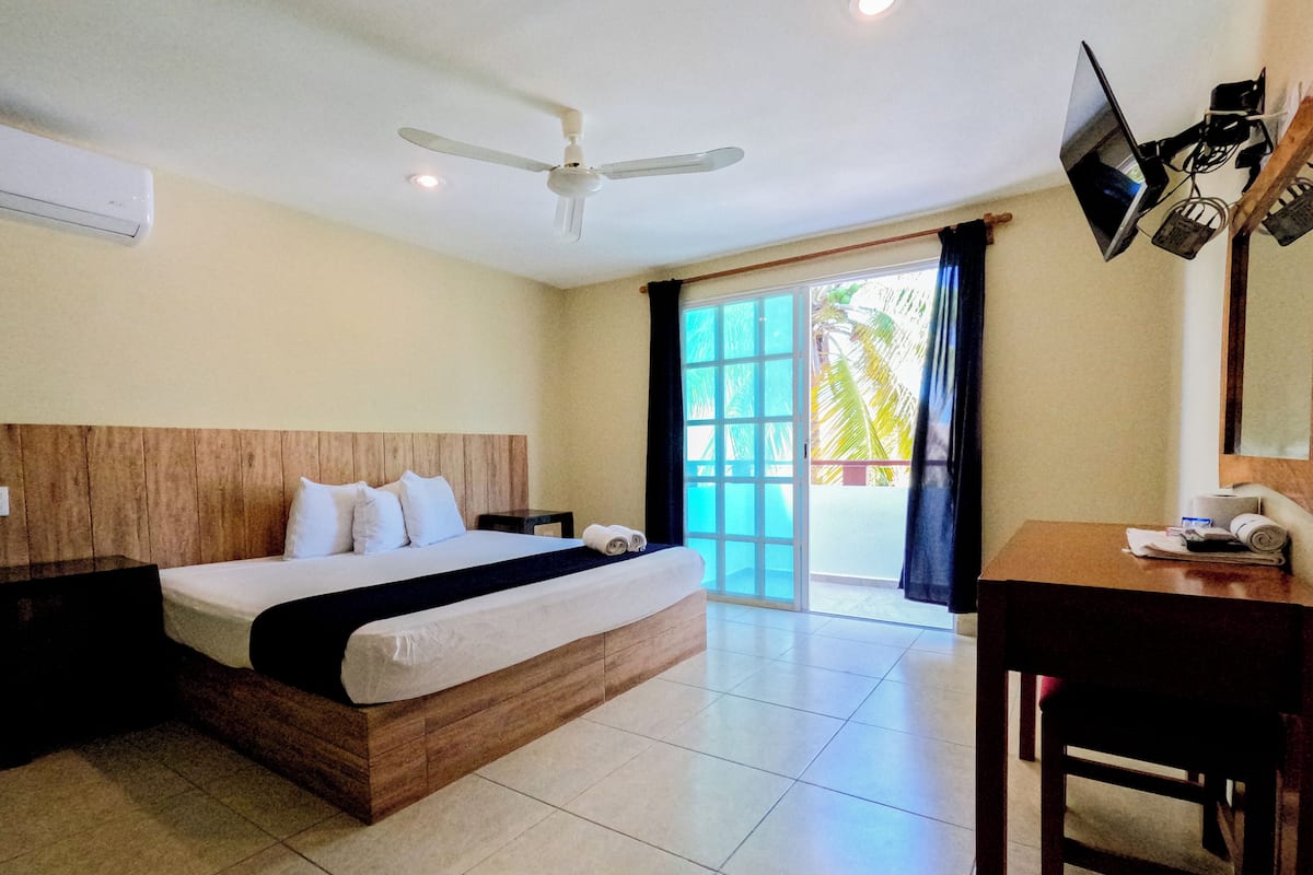 Quarto de hotel com uma cama, escrivaninha, ventilador de teto e TV fixada na parede.