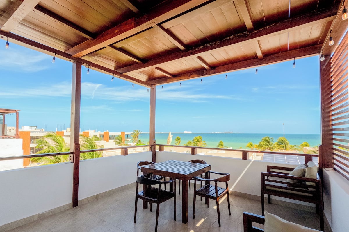Um espaço ao ar livre coberto com uma mesa e cadeiras, vista para o oceano e céu claro.