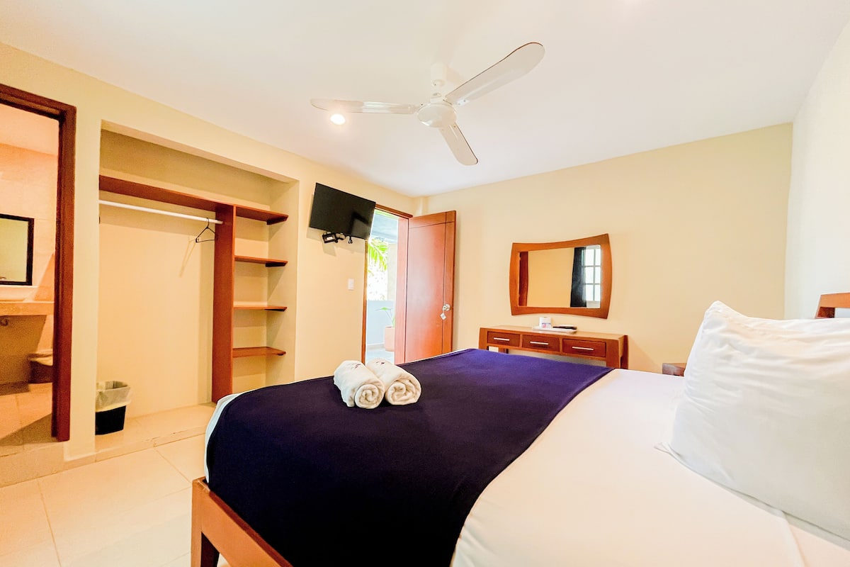 Quarto de hotel com cama, televisão, ventoinha no teto, cómoda de madeira e guarda-roupa.