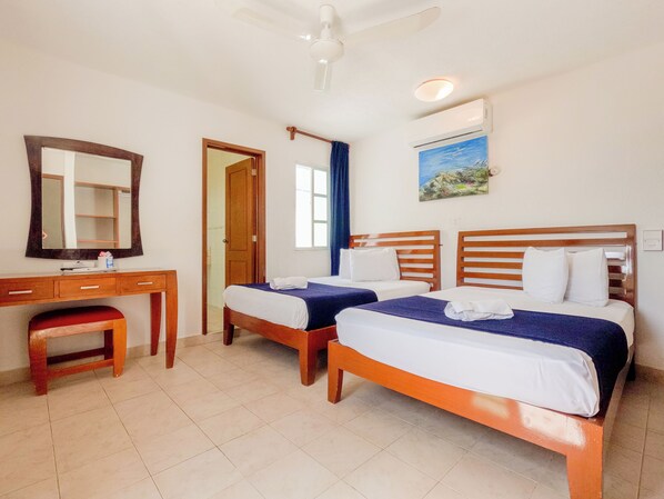 Quarto de hotel com duas camas de madeira, uma escrivaninha com espelho, um banquinho pequeno, um ventilador de teto e um quadro na parede.
