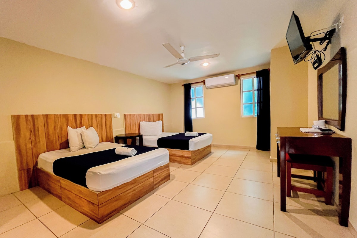 Quarto de hotel com duas camas, uma escrivaninha, uma cadeira, um ventilador de teto e um ar condicionado.