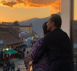 Um casal se abraçando em uma varanda com vista para uma rua com prédios e um pôr do sol.