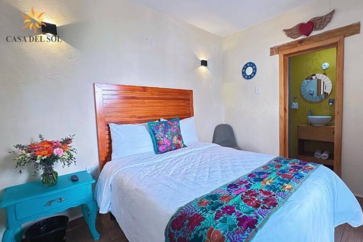 Um quarto com cabeceira de madeira, cama com duvet branco e colcha floral colorida, mesa de cabeceira turquesa com um vaso de flores e porta de madeira que dá para um banheiro com espelho redondo e pia.