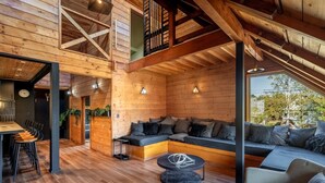 Um amplo quarto em estilo loft, com teto de madeira, área de estar integrada, mesa de jantar com cadeiras e uma grande janela com vista para o exterior.