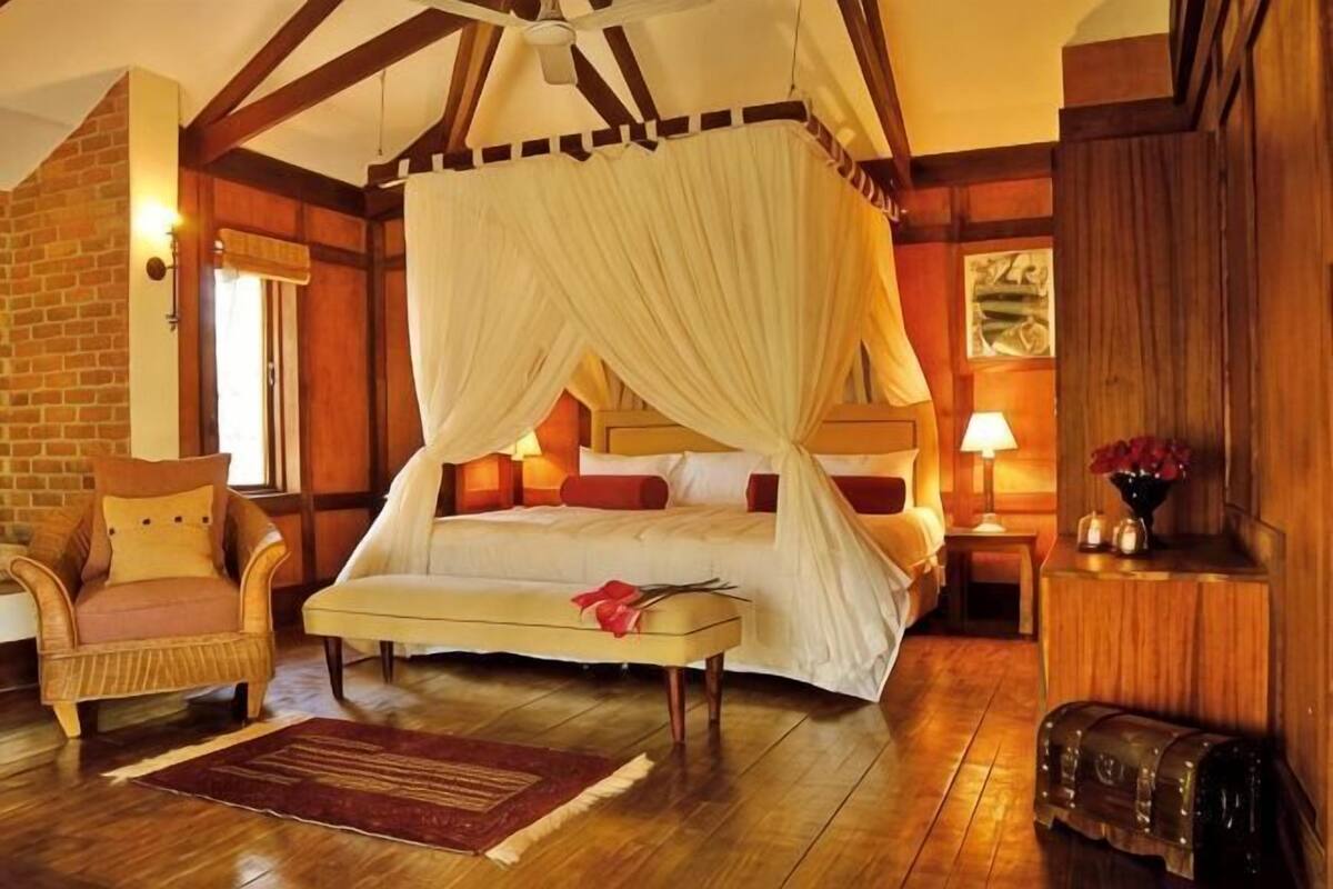Uma cama com dossel, um banco de madeira, uma mesinha de cabeceira com luminária e uma pequena mesa com um vaso.
