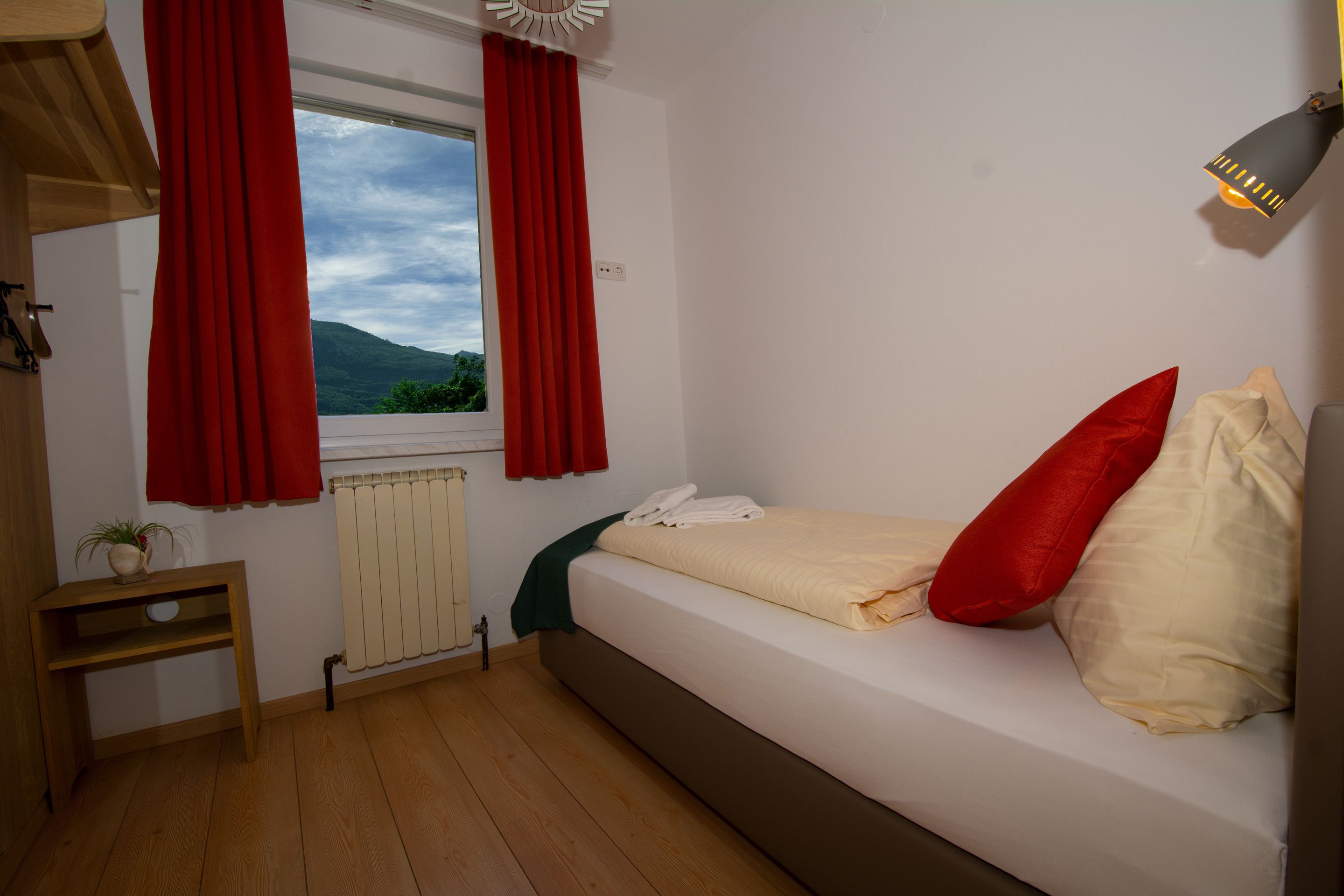 Quarto com cama, cortinas vermelhas, abajur, radiador e vista para o exterior.