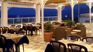Uma área de refeições ao ar livre com mesas e cadeiras, coberta por uma estrutura de madeira com treliça, e vista para o mar.