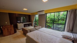 Quarto de hotel com uma cama grande, televisão, escrivaninha e janela com vista para o verde.