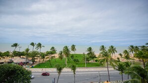 Vista da praia com palmeiras, uma estrada e a praia ao fundo.